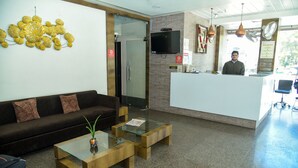 Uma área de recepção com um sofá, duas mesas com tampo de vidro, uma televisão e um balcão de atendimento.