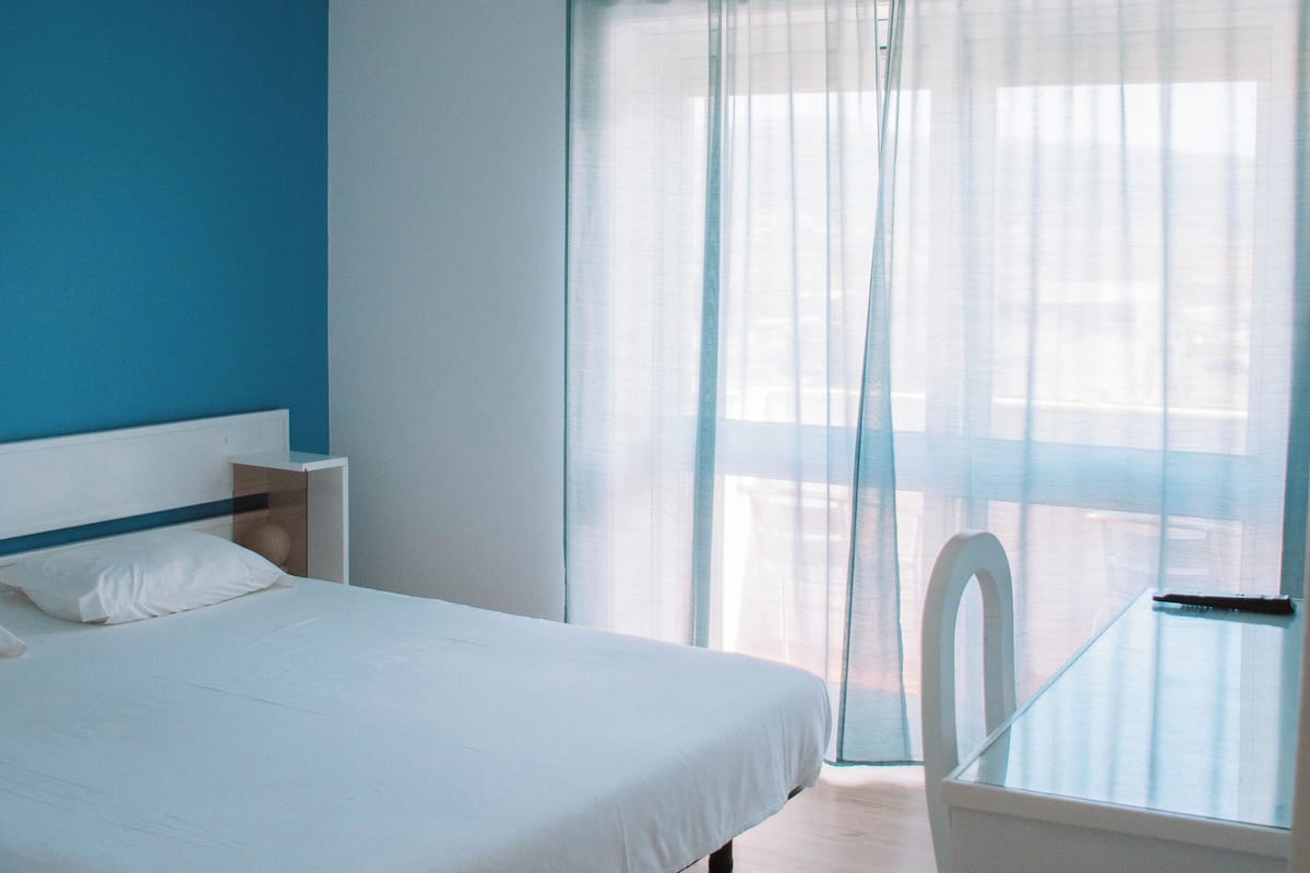 Quarto com cama, mesa de cabeceira e uma cadeira perto de uma janela com cortinas leves.