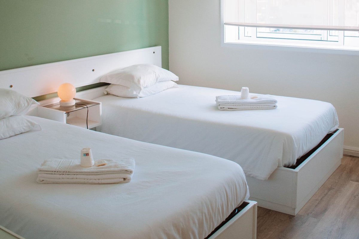 Quarto de hotel com duas camas, mesa de cabeceira e uma janela com persianas, e parede verde.