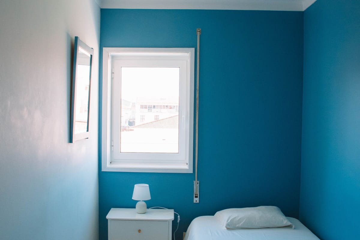 Quarto com uma cama de solteiro, uma mesinha de cabeceira, uma luminária e uma janela com vista para edifícios.