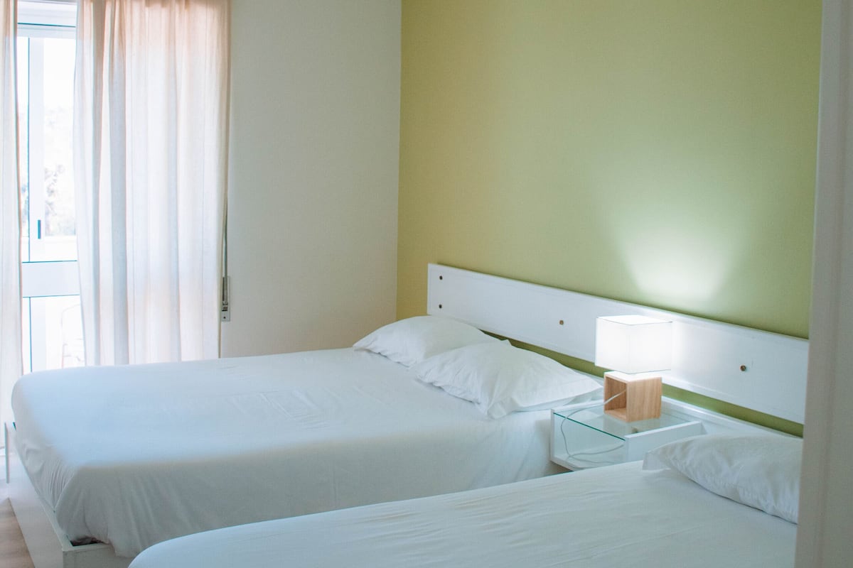 Quarto de hotel com duas camas de solteiro, mesa de cabeceira com abajur e janela com cortinas.