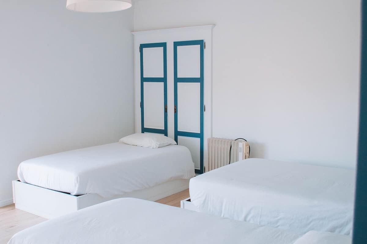 Quarto de hotel minimalista com duas camas de solteiro, uma luminária de teto branca e uma janela com persianas azuis.