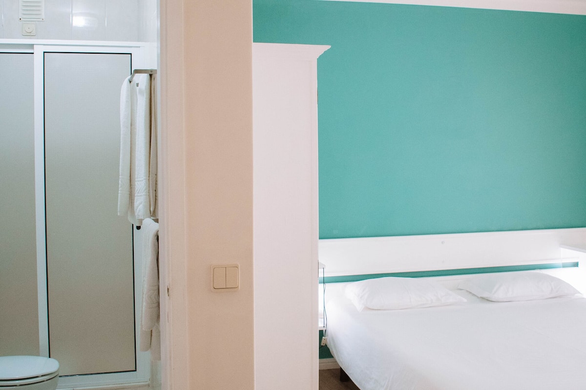 Quarto com cama, guarda-roupa branco, banheiro com vaso sanitário e chuveiro, e uma parede de destaque em tom verde-água.