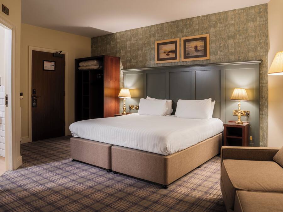 Quarto de hotel com uma cama grande, duas mesas de cabeceira com abajures, um guarda-roupa e um tapete xadrez.