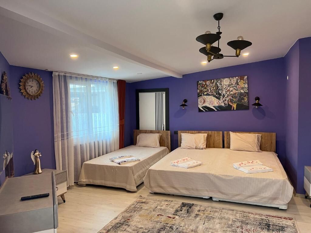 Quarto com duas camas, uma janela grande com cortinas, um ventilador de teto e um quadro na parede.