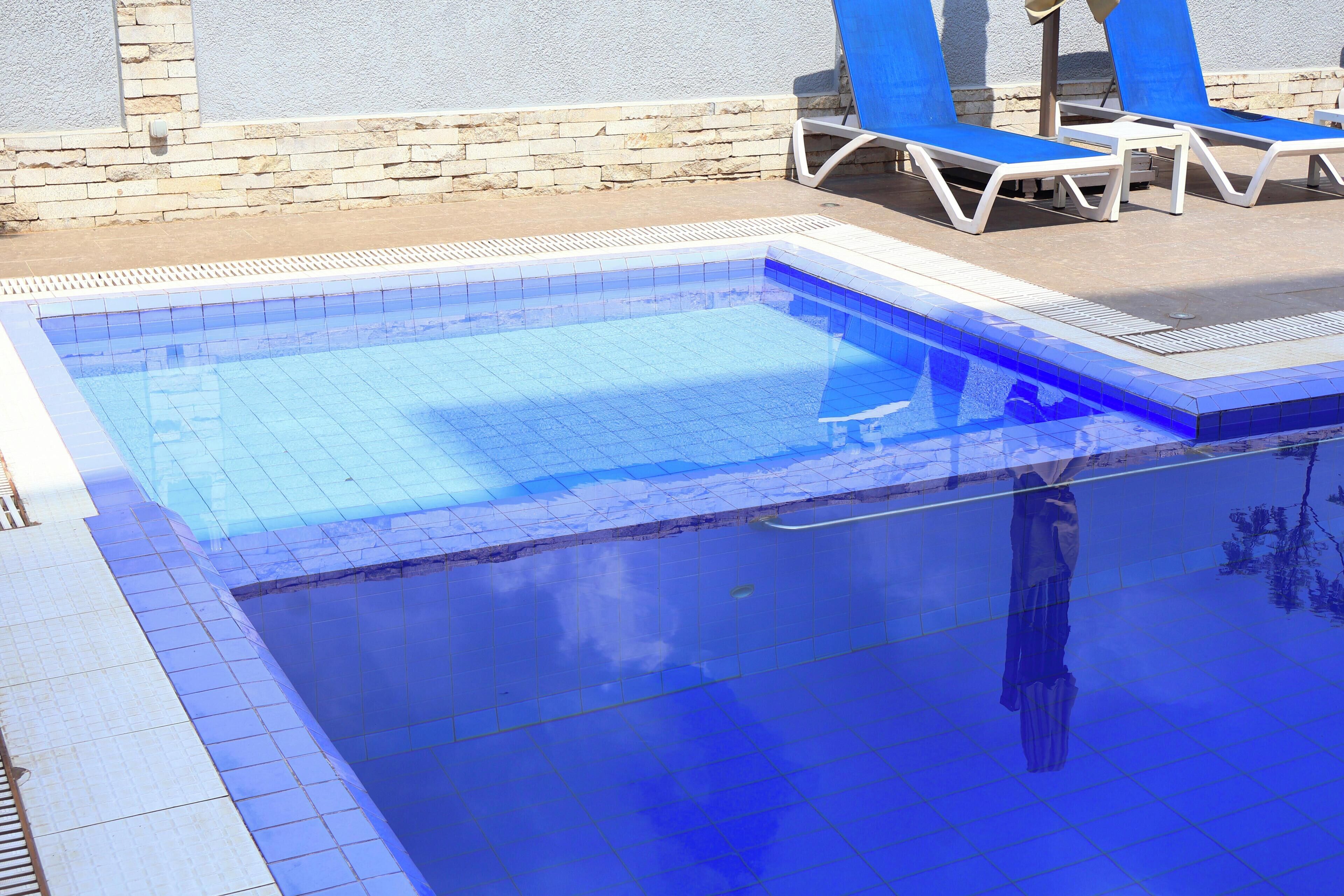 Piscina com azulejos azuis, parede de pedra e cadeiras de descanso brancas.