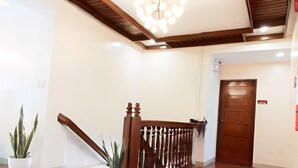 Um corredor com escada de madeira, paredes brancas e porta de madeira. Há duas plantas em vasos no chão.
