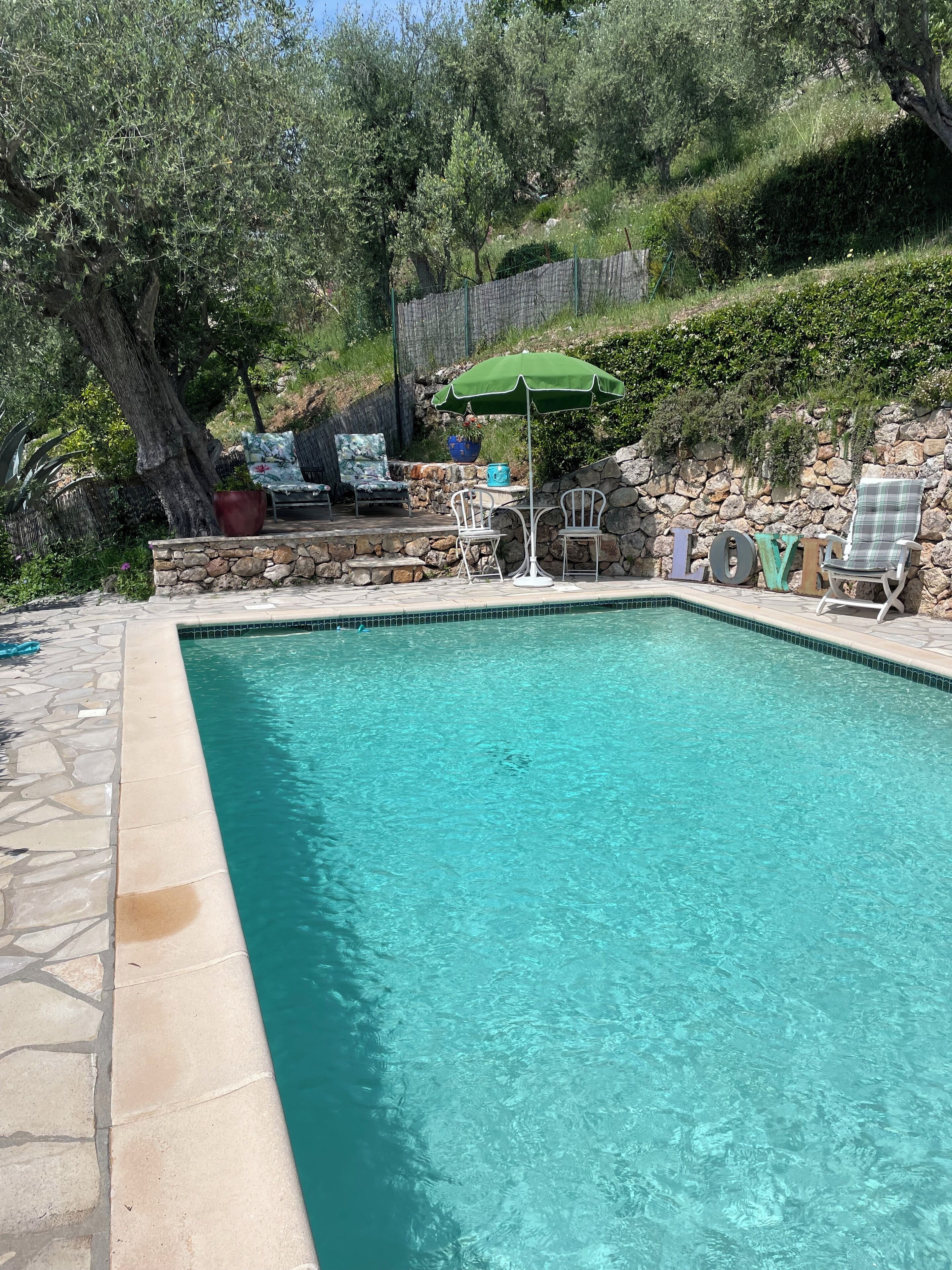 Piscina com azulejos de pedra, rodeada por um muro de pedra e uma área gramada com árvores e uma cerca.
