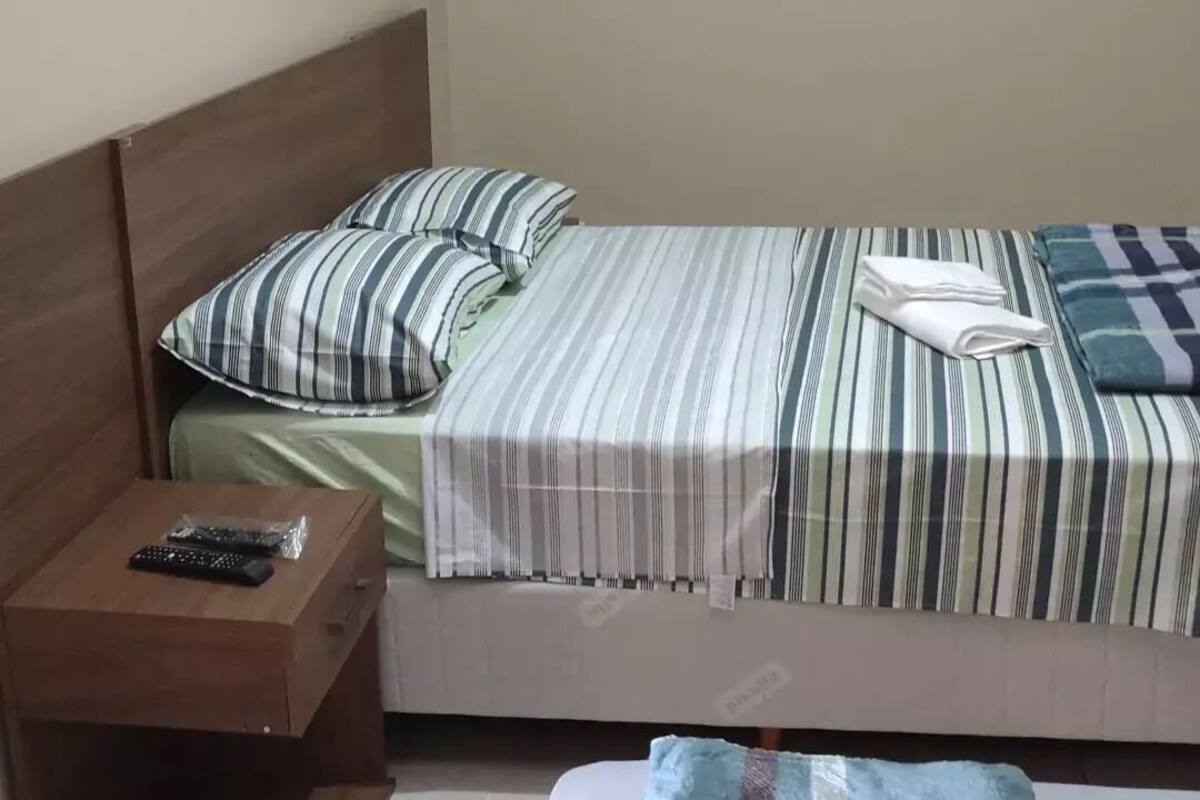Uma cama de solteiro com roupa de cama listrada, um criado-mudo de madeira com um controle remoto e um travesseiro azul e branco xadrez sobre um colchão branco.