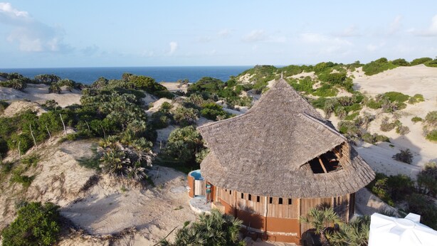 Construção com telhado de palha, com abertura central, rodeada por dunas de areia e vegetação rasteira, com vista para a área costeira.