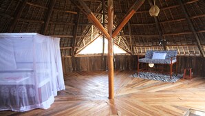 Quarto com piso de madeira, cama com mosquiteiro, mesa com almofada e telhado de palha.
