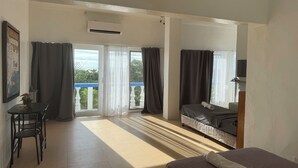Quarto com uma cama, uma escrivaninha e uma cadeira. Possui uma varanda com cortinas e vista para o verde.