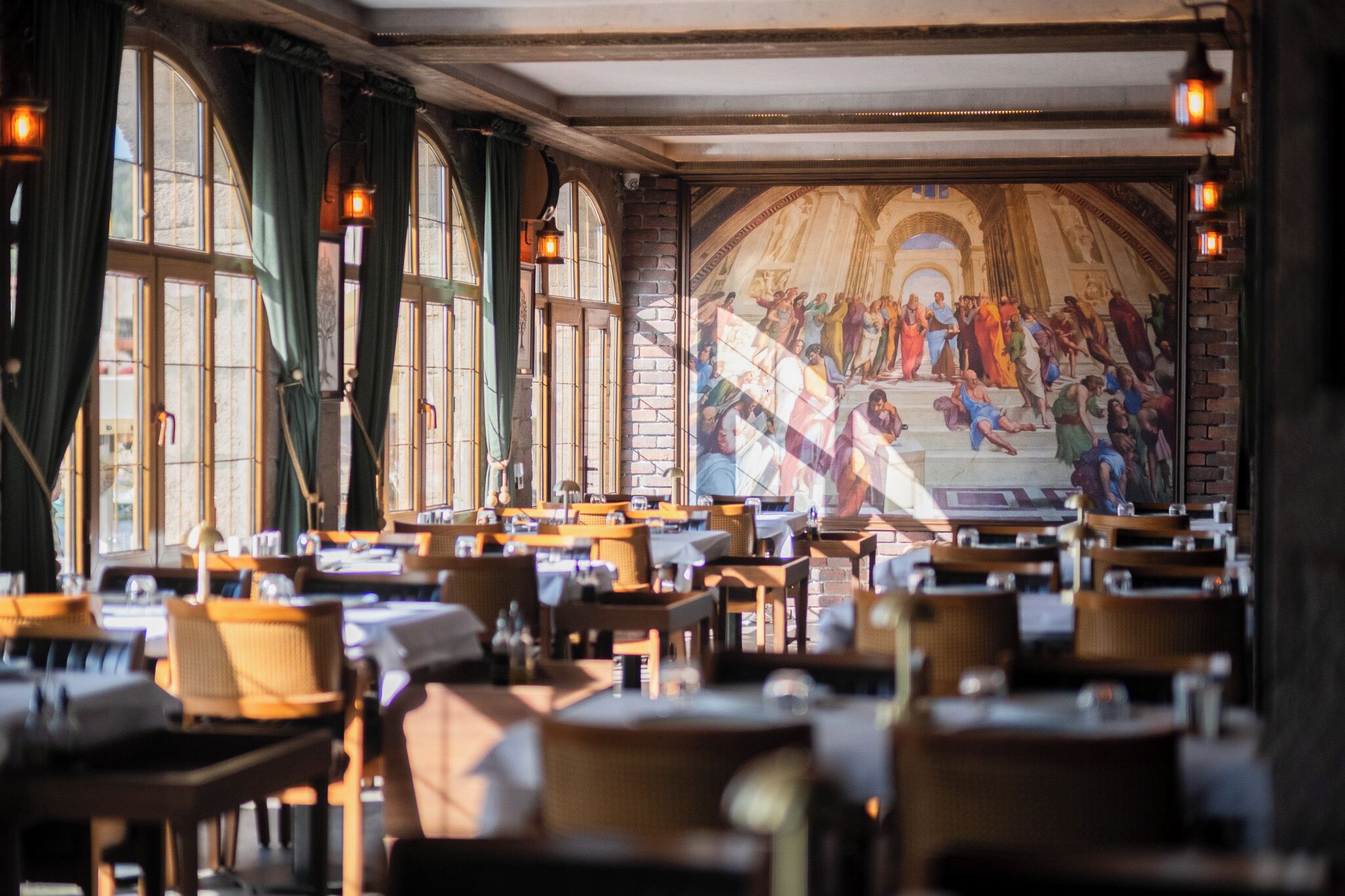Restaurante com pé-direito alto, grandes janelas e um mural retratando uma cena histórica. Mesas dispostas para refeições.