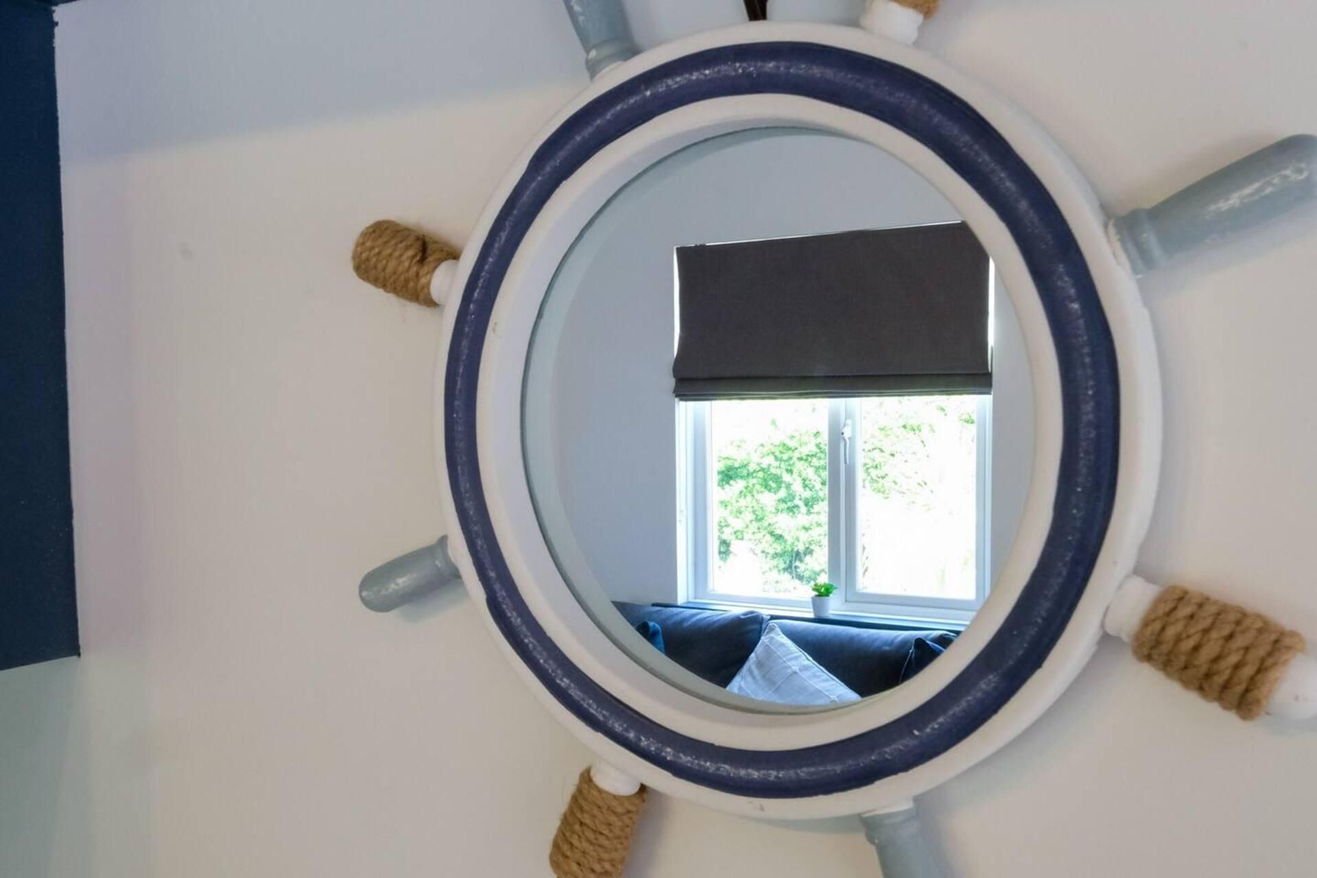 Espelho redondo com moldura escura, refletindo um quarto com janela, planta e almofada azul.
