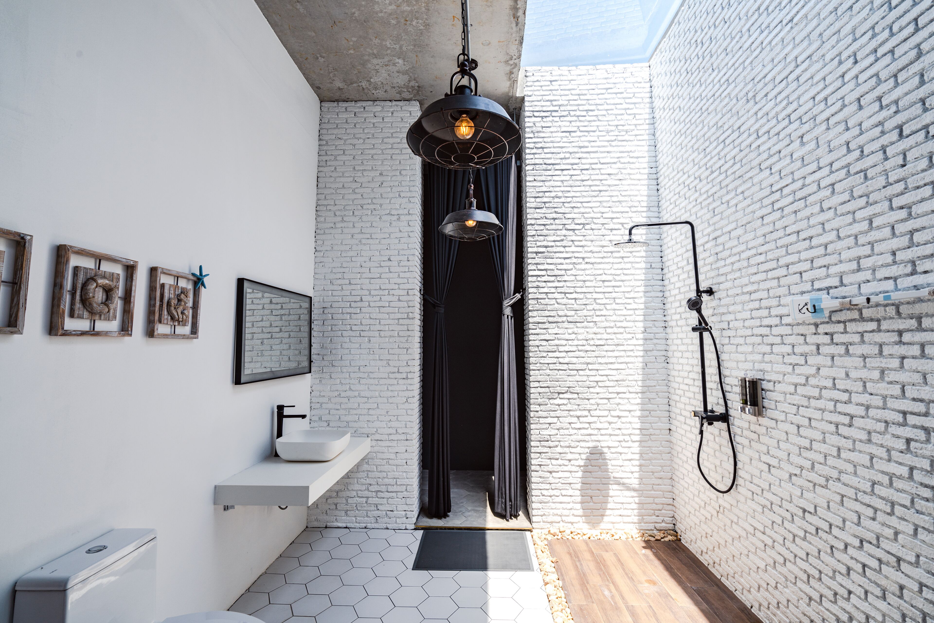 Casa de banho moderna com parede de tijolo branco, azulejos hexagonais, lavatório branco, sanita e chuveiro com cortina preta.
