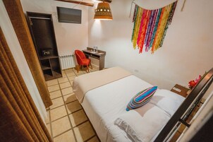 Quarto de hotel com cama, escrivaninha, cadeira, televisão e um painel de parede colorido.