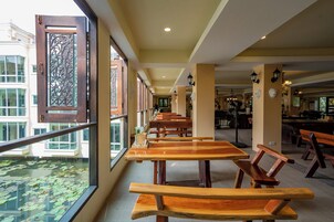 Um restaurante com mesas e bancos de madeira, grandes janelas com grades de metal decorativas e vista para um edifício com várias janelas.