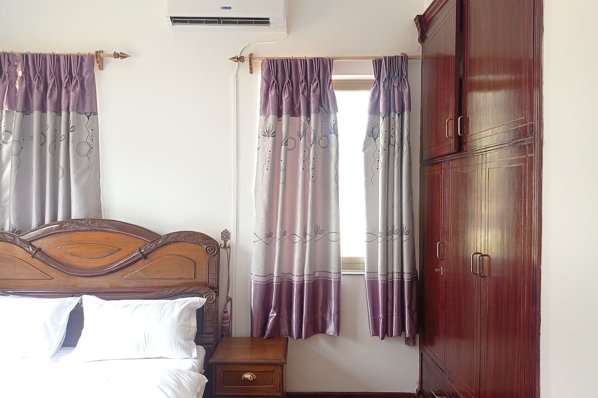Quarto com cabeceira de madeira, uma cama de solteiro com roupa de cama branca, mesa de cabeceira com gaveta e um guarda-roupa.