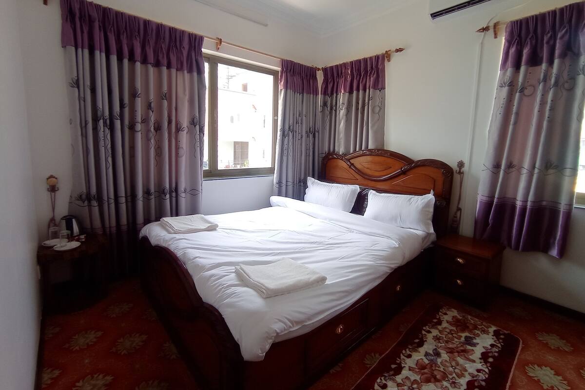 Cama bem arrumada com lençóis brancos, cabeceira de madeira e tapete vermelho estampado. Há uma mesa de cabeceira com abajur e uma pequena mesa com chaleira e xícaras.