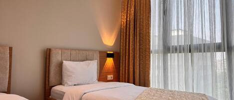 Quarto de hotel com duas camas, cabeceira com luminária e uma janela grande com cortinas leves.