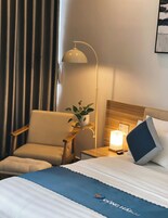 Quarto de hotel com cama, uma cadeira, mesa de cabeceira com luminária, planta e travesseiro.