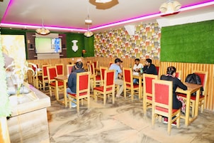 Restaurante com mesas de madeira e cadeiras vermelhas com almofadas, uma TV de tela plana na parede e um painel verde com padrão geométrico.