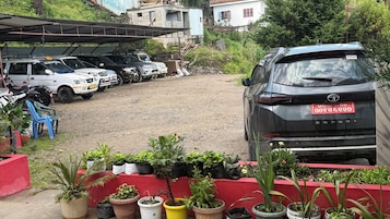Uma área de estacionamento com vários veículos, incluindo um SUV preto e uma van branca. Há plantas em vasos no primeiro plano e uma parede vermelha ao fundo.