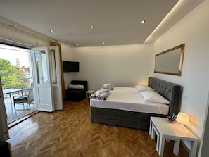 Quarto com cama grande, mesa de cabeceira, TV fixada na parede e varanda com mesa e cadeiras.