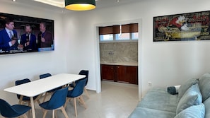 Uma sala de estar moderna com um sofá branco, uma área de jantar com mesa branca e cadeiras azuis, uma cozinha com painel de parede de mármore e uma TV exibindo um evento esportivo.