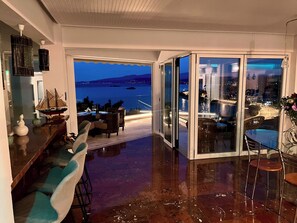 Uma sala de estar moderna com vista para o mar, uma área de jantar com mesa e cadeiras, e uma varanda envidraçada.