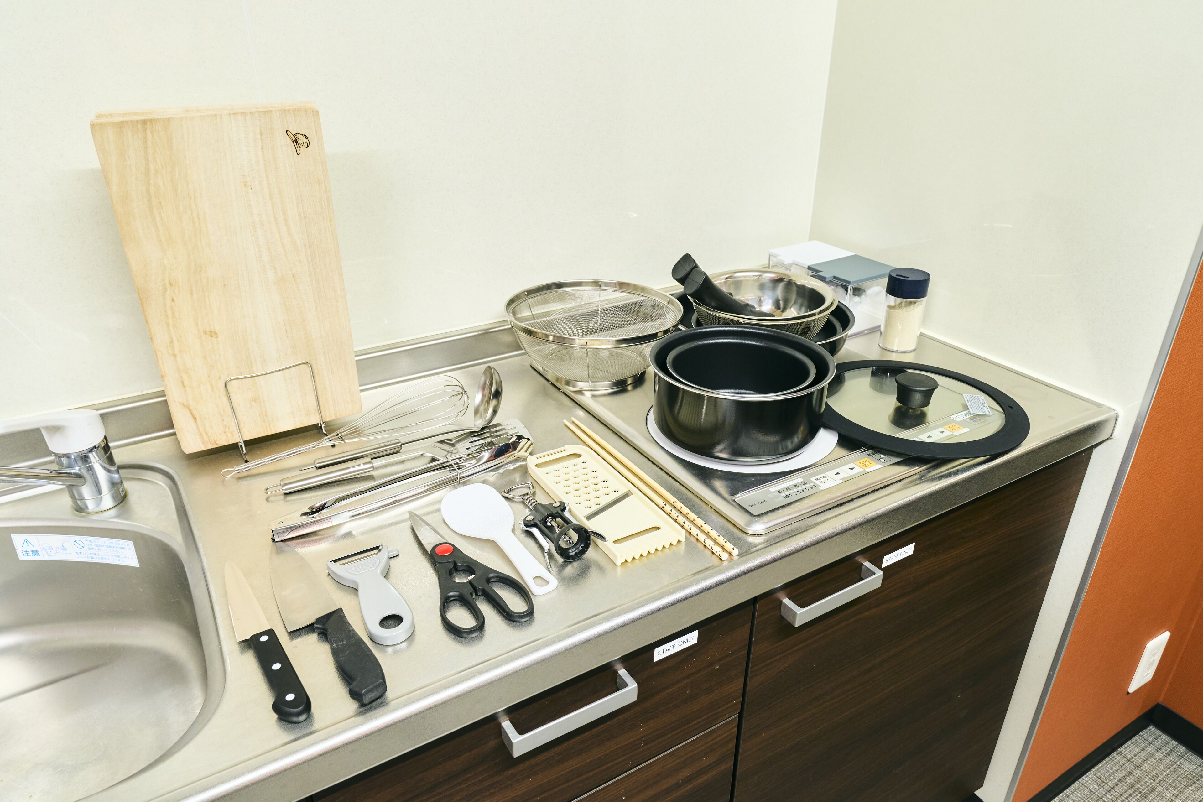 Uma bancada de cozinha com diversos utensílios, uma tábua de corte de madeira, uma panela, um escorredor e uma chaleira.