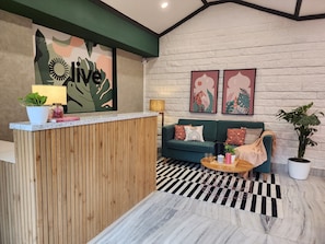 Uma área de recepção com balcão de madeira, um sofá verde com almofadas estampadas e um tapete quadriculado.