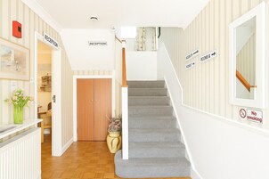 Uma área de recepção com escadas que levam a um segundo andar, um aviso de 'Proibido Fumar' e placas indicativas para 'Quarto 19' e 'Quarto 20'.