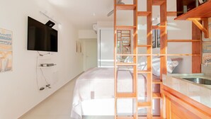 Quarto com cama, televisão na parede, estante de madeira e área de cozinha com pia.