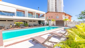 Piscina com espreguiçadeiras, área de restaurante e um prédio alto ao fundo.