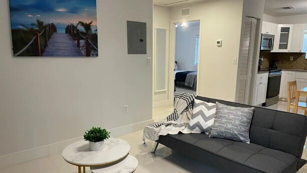 Uma sala de estar moderna com um sofá, uma mesinha redonda com uma planta e um quadro de uma paisagem praiana.