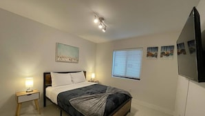 Quarto com cama, abajures, iluminação no teto, janela com persianas e arte nas paredes.