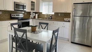Uma cozinha moderna com armários brancos, geladeira de aço inoxidável, micro-ondas e forno. Há uma mesa de jantar com cadeiras e uma orquídea em vaso.