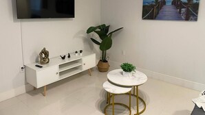 Uma sala de estar moderna com uma TV de tela plana, um console branco com objetos decorativos, uma planta verde grande e uma mesa redonda com moldura dourada.
