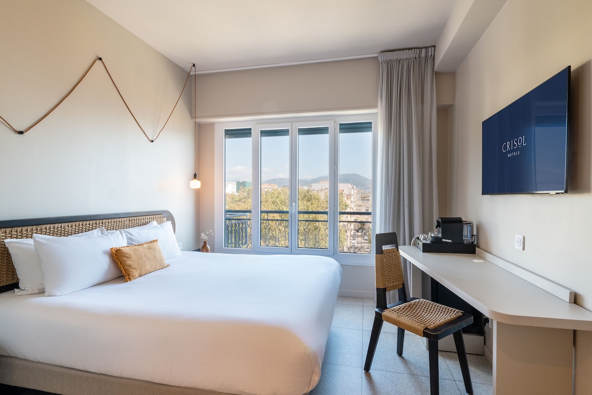 Quarto de hotel com cama, escrivaninha, cafeteira, televisão e janela com vista para edifícios e colinas.
