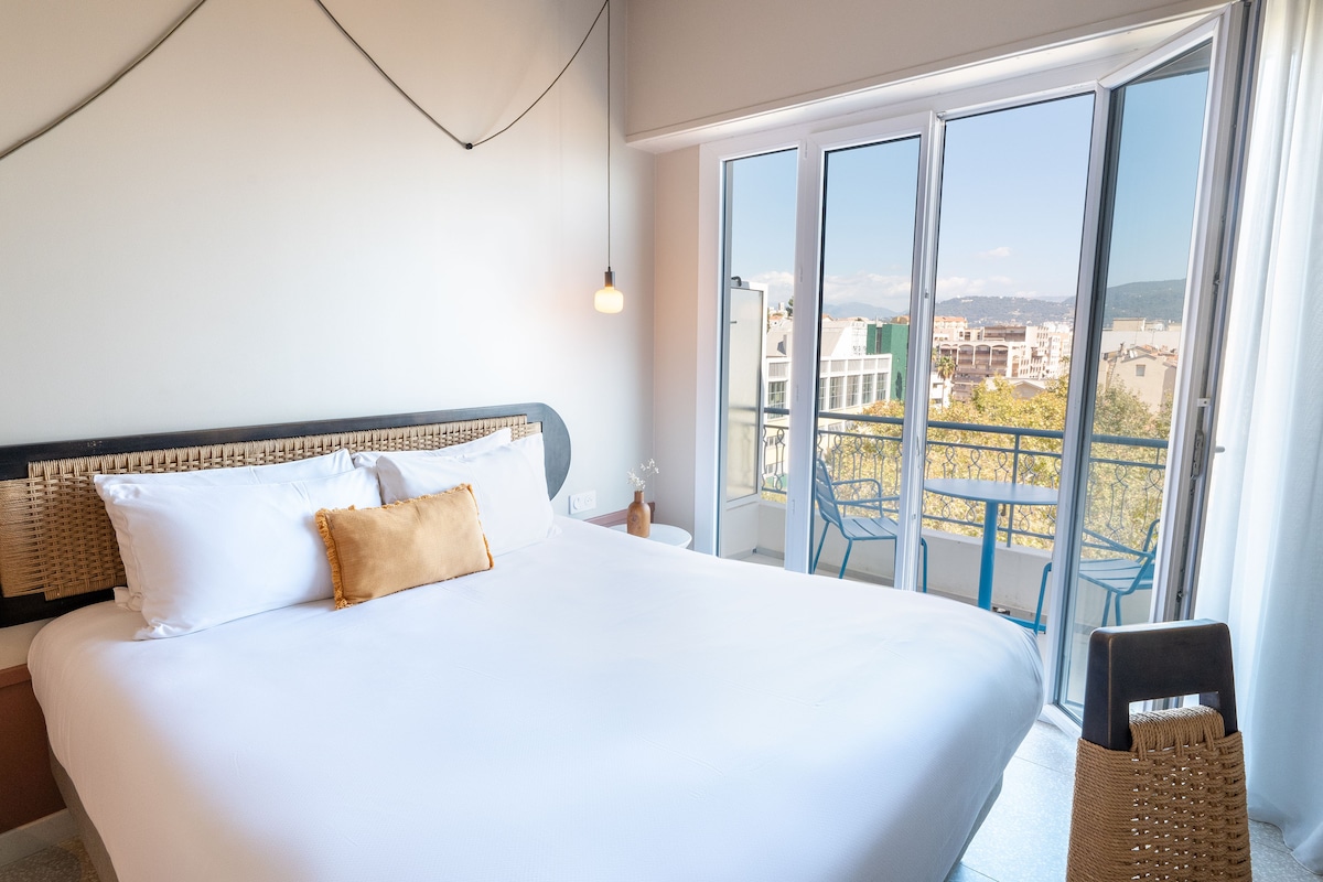 Quarto de hotel com uma cama grande, vista da cidade através de janelas do chão ao teto e varanda com cadeiras azuis.
