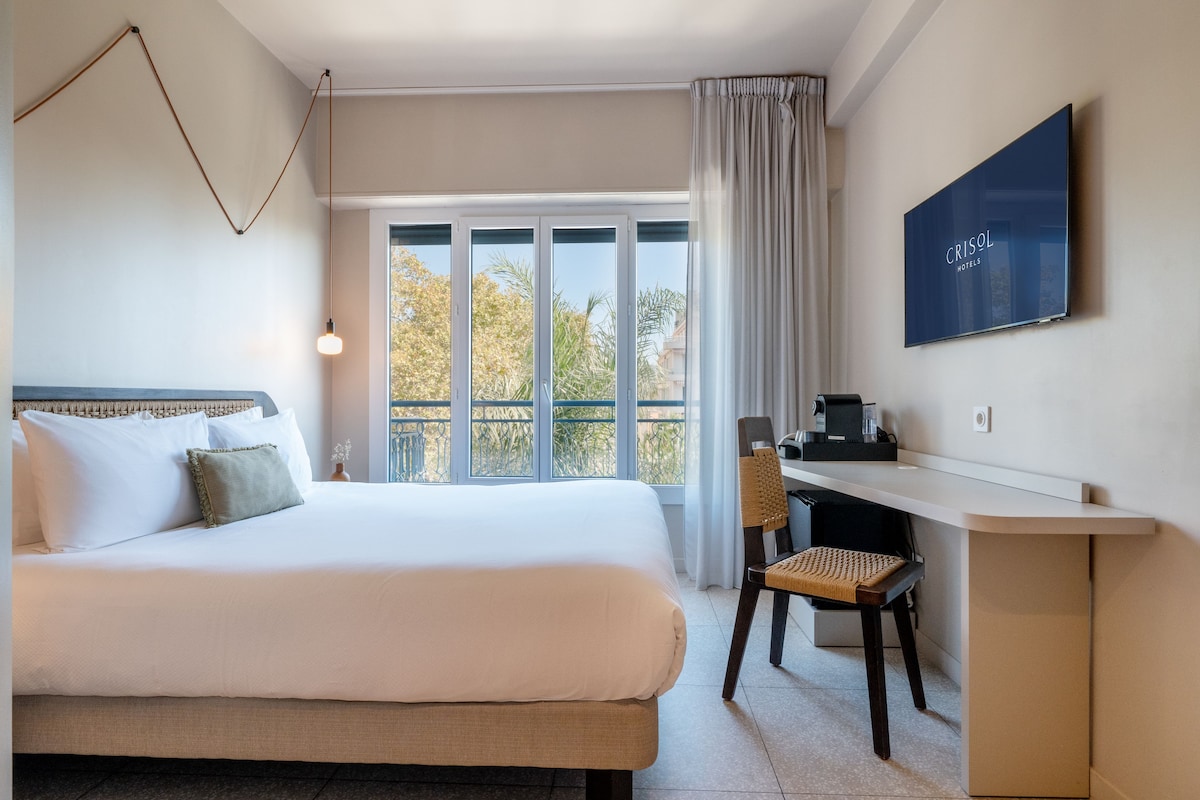 Quarto de hotel com cama, escrivaninha, cadeira, televisão e janela com vista para árvores.