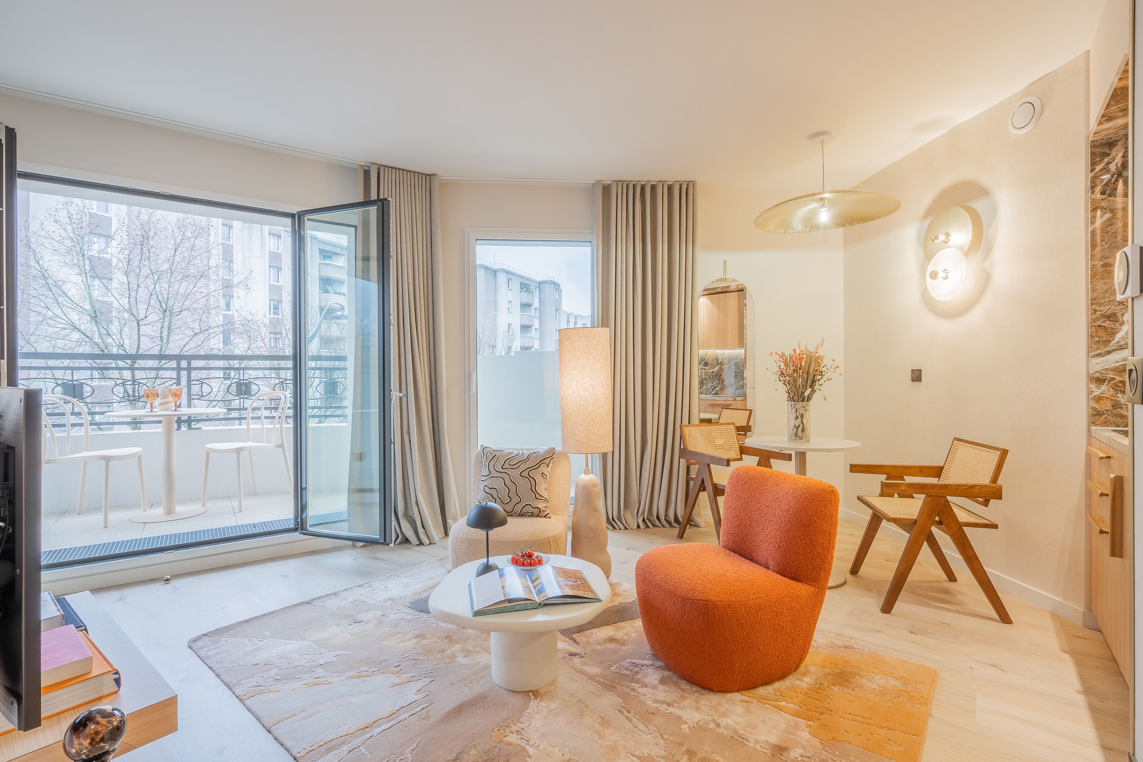 Uma sala de estar moderna com uma grande porta de vidro que dá para um terraço ao ar livre, uma mesa de centro redonda com um livro, uma poltrona laranja e uma área de jantar com cadeiras de madeira.