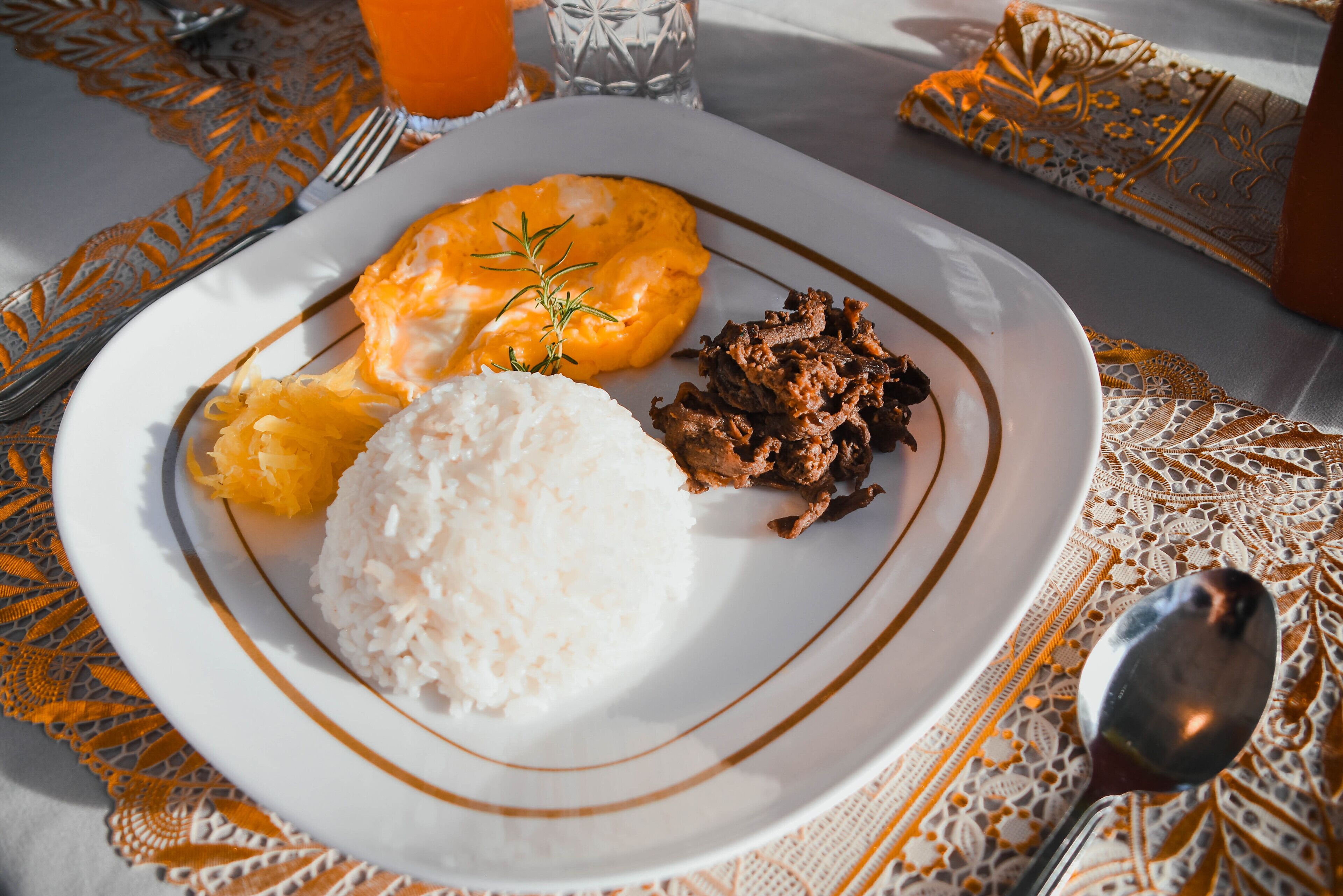 Prato de comida com arroz, um acompanhamento amarelo e um prato com carne marrom, servido em prato branco com borda dourada.