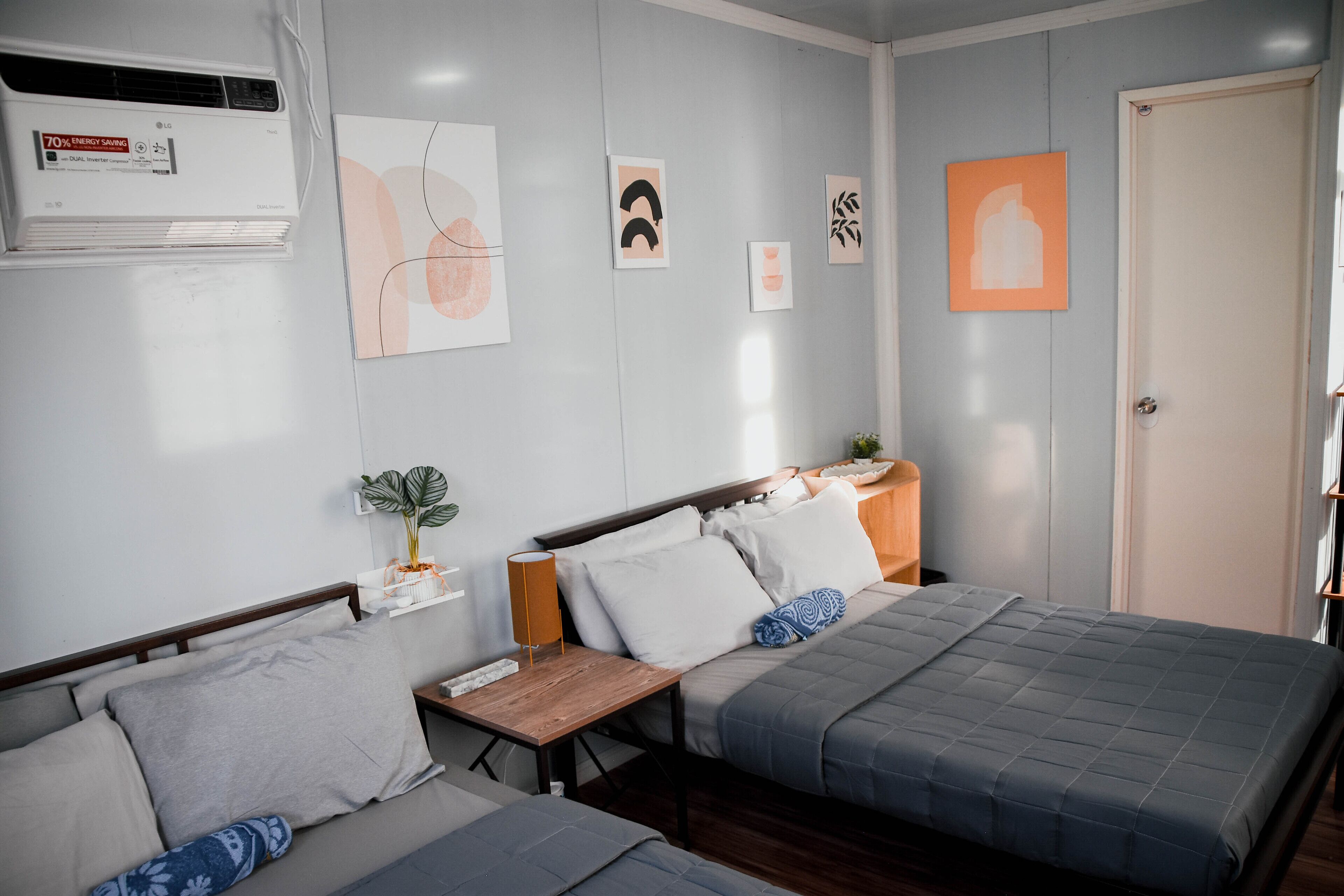 Quarto de hotel com duas camas, uma mesinha de cabeceira com abajur, uma pequena planta e uma obra de arte na parede.