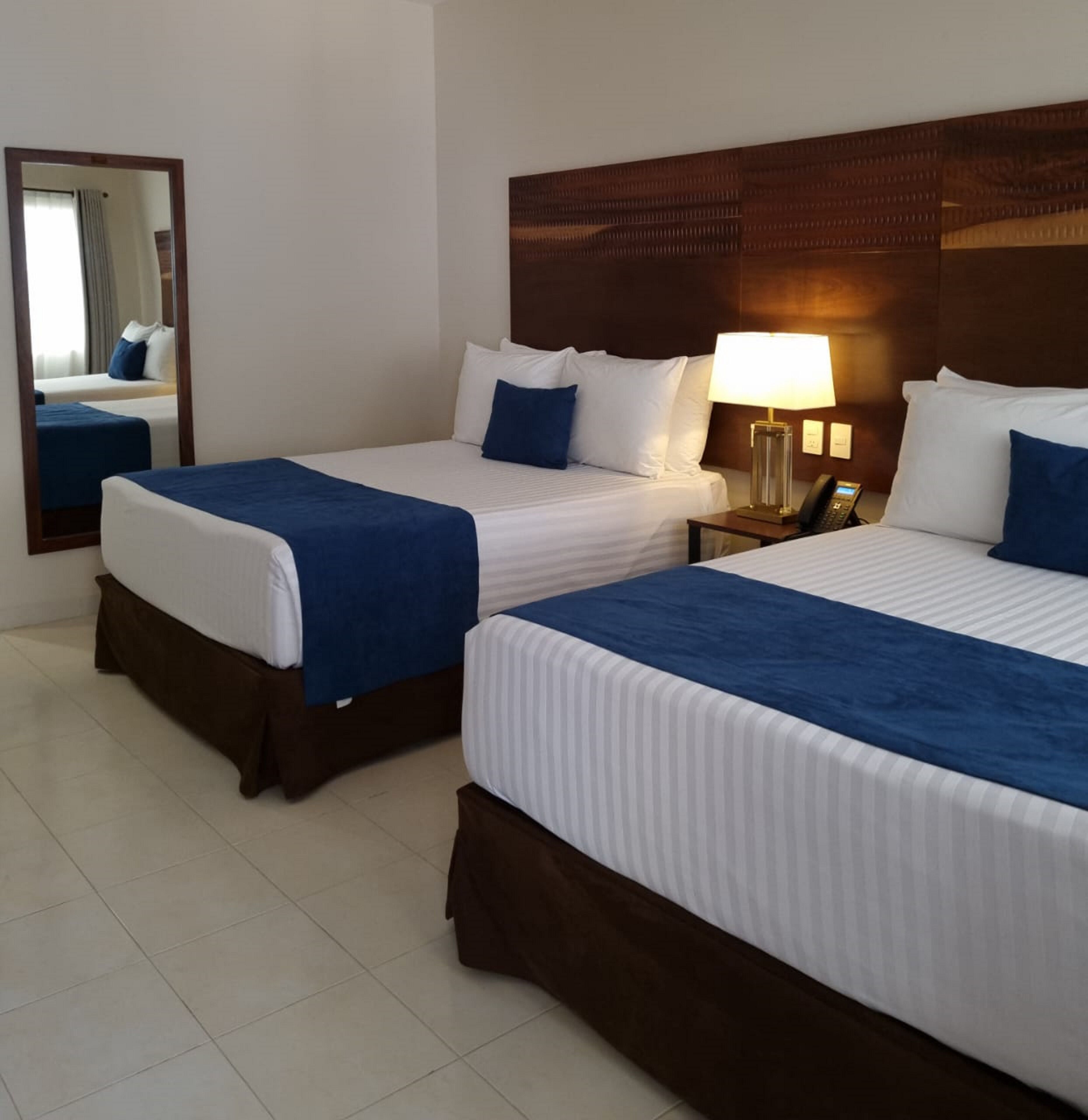 Quarto de hotel com duas camas, cada uma com colcha azul e travesseiros brancos. Há uma mesa de cabeceira com abajur e telefone.