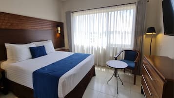 Quarto de hotel com uma cama grande, uma escrivaninha, uma cadeira, uma televisão e uma janela com cortinas.