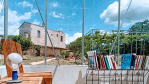Quarto com uma grande janela que dá para um edifício de pedra e vegetação. Há uma mesa com um abajur e uma estante de livros.