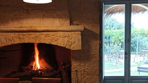 Lareira de pedra com fogo visível, mesa com cadeiras e porta que dá para o exterior.
