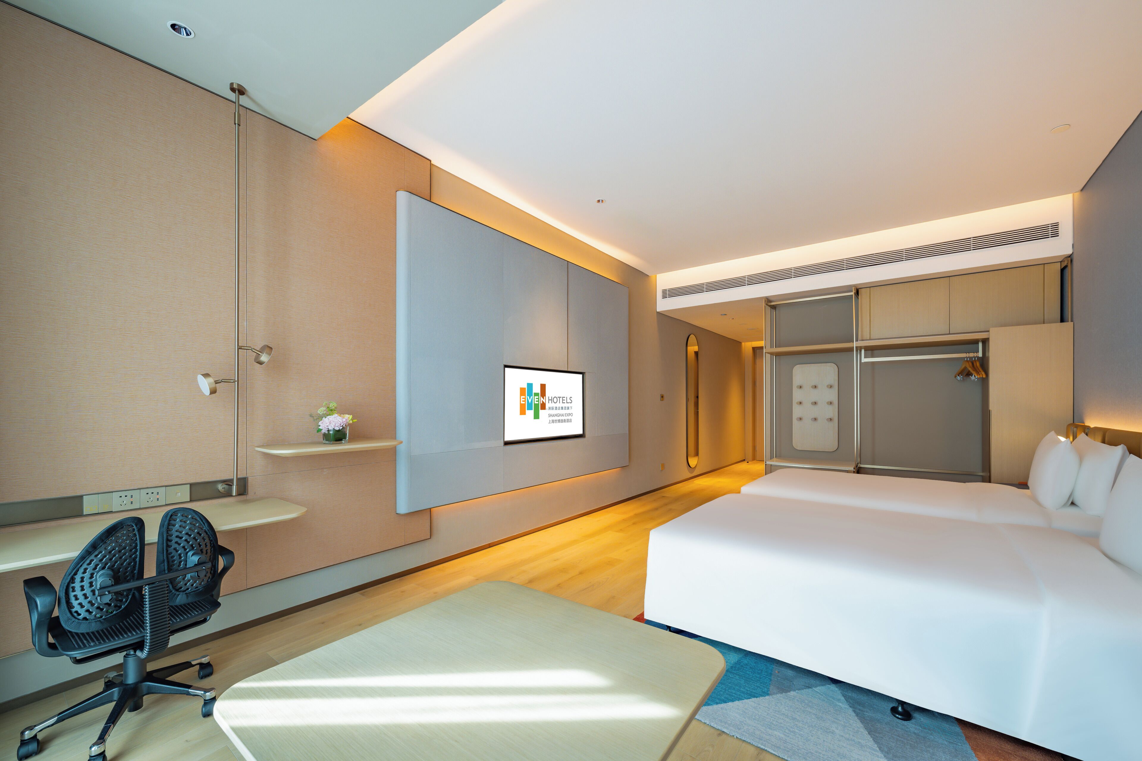 Quarto de hotel com cama, escrivaninha, cadeira, guarda-roupa e um pequeno prateleira com uma flor.